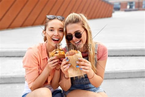 休闲、快餐和友谊的概念 快乐微笑的少女或戴墨镜的好朋友在夏天在城市街道上吃汉堡。高清摄影大图 千库网