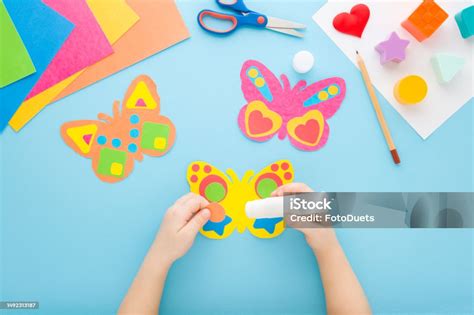 어린 아이의 손은 접착제 막대기를 들고 파란색 테이블 배경에 있는 종이에서 화려한 나비 모양을 붙인다 파스텔 색상 장식 요소 만들기 근접 촬영 시점 촬영 하향식 보기 나비에