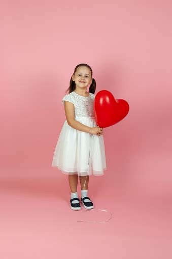 분홍색 배경에 고립 된 빨간 하트 모양의 풍선을 들고 흰색 드레스를 입은 전체 길이의 부주의하고 무관심한 소녀 2월에 대한 스톡