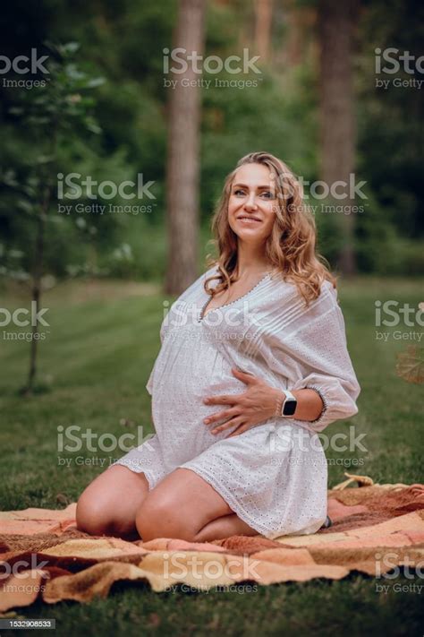 하얀 보헤미안 여름 드레스를 입은 임산부는 숲 배경 근처에서 임신한 배에 손을 얹고 담요 위에 앉아 화창한 날에 귀여운 미소를