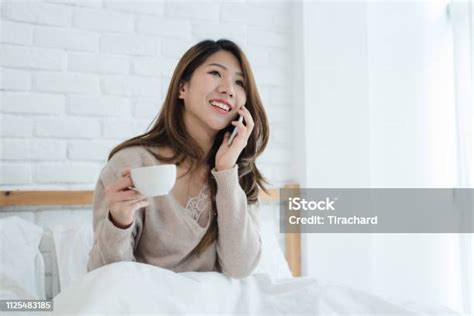 아시아 여자는 아침에 커피 한잔 들고 하는 동안 그녀의 침대에는 스마트폰을 사용 하 여 아름 다운 아시아 여자 즐기는 따뜻한