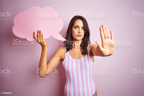스트라이프 핑크 수영복을 입고 진지하고 자신감있는 표정으로 정지 표지판을하는 열린 손으로 분홍색 배경 위에 말하는 풍선을 들고