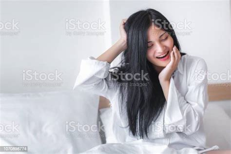 아름 다운 젊은 아시아 여자는 일어나서 흰 침대에서 아침에 흰 셔츠와 함께 행복 한 미소를 만들 가정의 방에 대한 스톡 사진 및