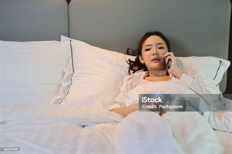하얀 잠옷을 입은 젊고 아름다운 아시아 여성이 잠자리에 들기 전에 모바일 스마트폰이 있는 침실에 누워 좋은 꿈을 꾸고 있다