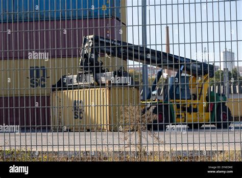 Reach Stacker Lifting A Shipping Container Behind A Wire Mesh Fence In