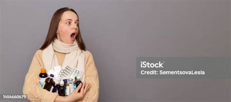 놀란 백인 여성의 초상화는 점퍼와 스카프를 입고 알약을 들고 광고 영역을 바라보며 놀라서 비명을 지르며 회색 배경 위에 고립되어 서 있습니다 30 34세에 대한 스톡 사진