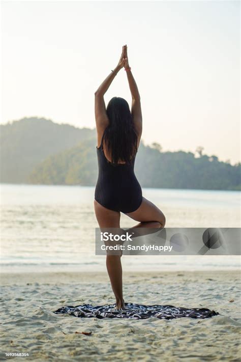 한 여자가 한쪽 다리에 수영복을 입고 고요한 해변에서 나무 요가 포즈를 연습하는 동안 태국의 여름 방학 동안 일몰에 촬영한 전체 길이의 뒷모습 여자에 대한 스톡 사진 및