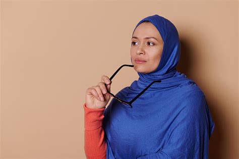 青いヒジャーブを着た自信に満ちたアラブのイスラム教徒の女性眼鏡を持ちベージュの背景に思慮深く目をそらす 1人のストックフォトや画像を多数ご