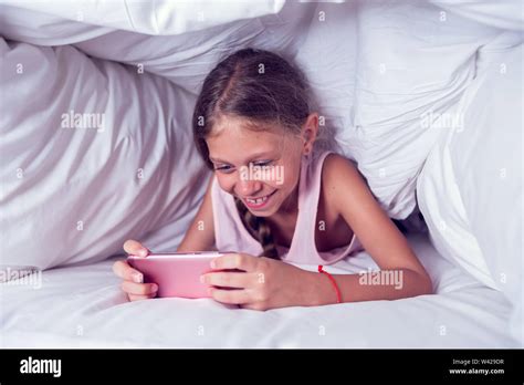 Happy Girl Looking At Mobile Phone While Lying Under Blanket On The Bed