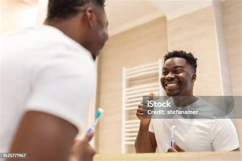 칫솔을 들고 미소를 짓고 욕실에서 거울을 보고 있는 아프리카 남자 남자는 화장실에서 아침에 자신의 이빨을 닦고 잠옷에 잠자는