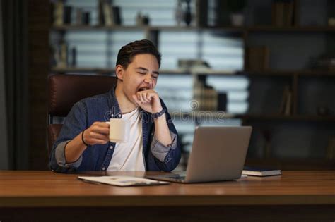 Sleepy Employee Sitting At Dark Office Using Laptop Drinking Coffee