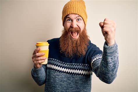 빨간 머리 아일랜드 남자 수염 을 입고 겨울 모자 마시는 떨어져 뜨거운 커피 자랑 스럽게 비명을 지르며 승리와 성공을 축하 매우 흥분 응원 감정 감정에 대한 스톡 사진 및