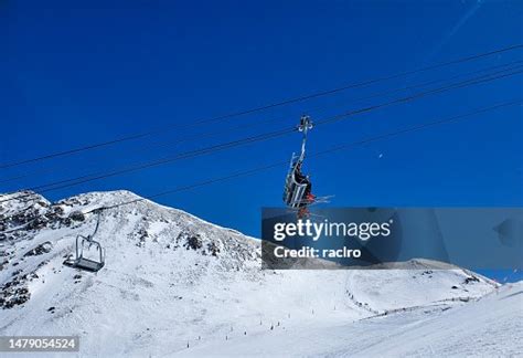 Looking Up At Skiers On A Ski Lift With A Towering Peak And Blue Sky