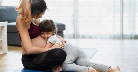 아시아의 젊은 사랑스러운 딸이 아름다운 임산부 배를 껴안고 있다 매력적인 임신 예정 소녀는 집에서 거실에서 요가 운동을 하는 동안 행복과 사랑으로 어린 소녀를 포용합니다