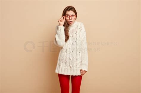 尴尬高清图片 尴尬的年轻女模特手放在眼镜边缘，表情困惑，惊讶地发现一些令人难以置信和难以置信的东西，穿着冬季毛衣，隔离在米色墙上 产品实拍免费