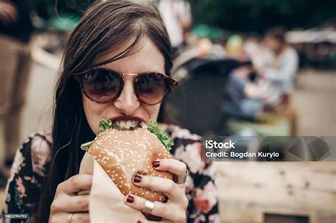 수 분이 많은 햄버거를 먹는 Hipster 세련 된 여자 Boho 소녀 맛 있는 치즈 버거를 물고 길거리 음식 축제에 웃 고 여름입니다 여름 휴가 여행 피크닉 텍스트를 위한