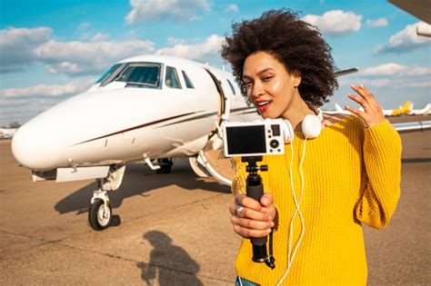 젊은 여성 동영상 블로거는 자신을 촬영 그녀의 카메라로 셀 카 스틱에 장착 된 공항 활주로에 주차 된 개인 비행기 옆에 Monopod에 대한 스톡 사진 및 기타 이미지