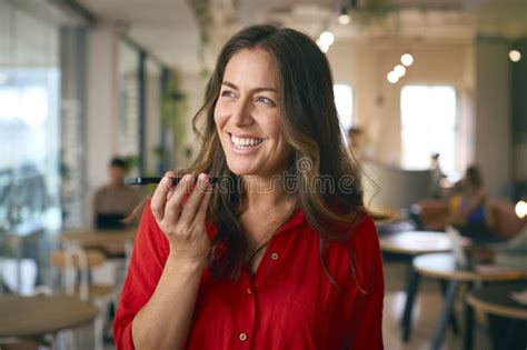 Smiling Mature Businesswoman Standing In Busy Office Talking Into Mic