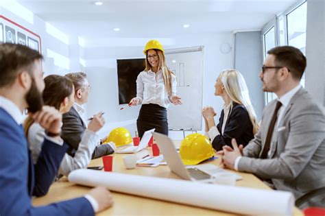 젊은 매력적인 성공적인 백인 여성 건축가 공식적인 착용을 입고 머리에 노란 헬멧 화이트 보드와 얘기 앞에 서 그녀의 동료는 앉아서 듣고 모던 양식에 대한 스톡 사진 및 기타