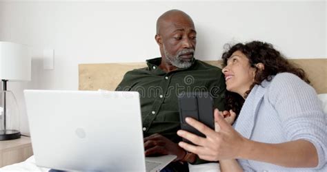 Happy Mature Diverse Couple Sitting On Bed Using Laptop And Smartphone