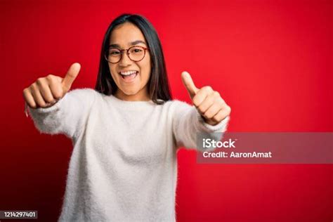 젊은 아름 다운 아시아 여자 캐주얼 스웨터와 안경을 입고 빨간 배경 에 긍정적 인 제스처를 수행 하는 손 엄지 손가락 미소와