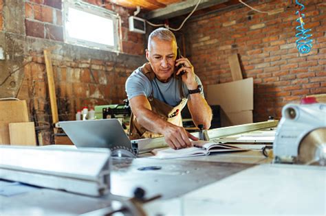 목공 가게에서 노트북의 목록을 확인하는 동안 휴대 전화를 사용하는 성숙한 목수 실제 인물에 대한 스톡 사진 및 기타 이미지