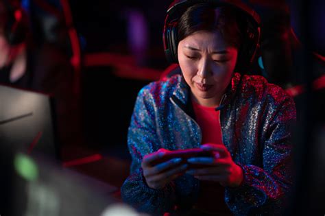 귀여운 젊은 아시아 소녀는 열정적으로 컴퓨터 클럽에서 그녀의 스마트 폰에서 재생 그녀의 친구는 서로 온라인 게임을하는 동안 Brand Name Video Game에 대한