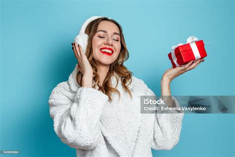 하얀 가짜 모피 코트를 입은 젊은 행복한 여성은 미소를 짓고 웃고 춤을 추고 음악을 듣고 스튜디오의 푸른 배경에 고립 된 손에 빨간 선물 상자를 들고 있습니다 12월에 대한