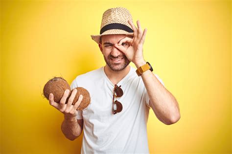 여름 모자를 쓰고 노란 배경 위에 코코넛 과일을 들고 행복한 얼굴로 손가락을 통해 보는 눈으로 확인 사인을하고 미소 짓는 젊은