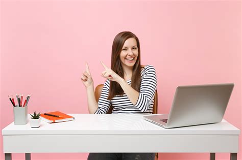 젊은 웃는 여자 캐주얼 옷 지점 검지 손가락 옆으로 앉아 파스텔 핑크 배경에 고립의 현대 Pc 노트북과 흰색 책상에서 작동