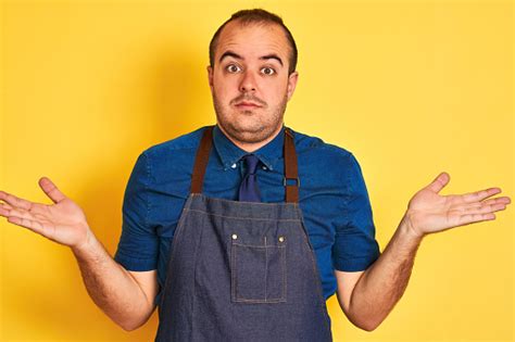 고립된 노란색 배경 위에 서있는 앞치마를 입은 젊은 상점 주인 남자는 팔과 손을 들고 혼란스러운 표정을 짓고 있습니다 의심 개념 남성에 대한 스톡 사진 및 기타 이미지