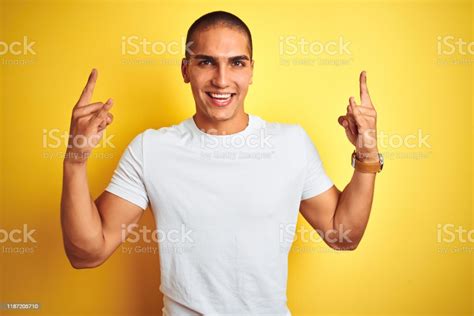 노란색 고립 된 배경 위에 캐주얼 한 흰색 티셔츠를 입은 젊은 백인 남성이 미친 표정으로 손을 들고 바위 기호를하고 있습니다 음악 스타 무거운 개념 T 셔츠에 대한 스톡