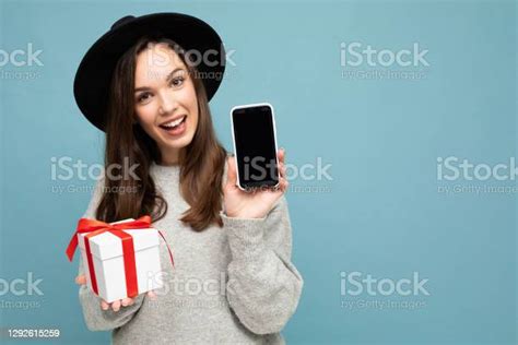 세련된 검은 모자와 회색 스웨터를 입고 파란색 배경 벽에 고립 된 예쁜 웃는 긍정적 인 젊은 갈색 머리 여성의 사진은 모형을위한 스마트 폰 화면 디스플레이를 보여주고