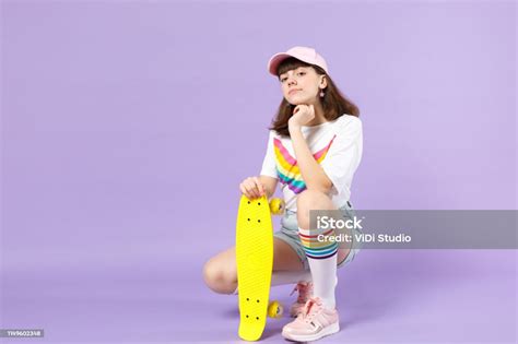 노란 스케이트 보드를 들고 앉아 생생한 옷에 귀여운 십 대 소녀 보라색 파스텔 벽 배경에 고립 된 턱에 손을 소품을 넣어 사람들은 진심으로 감정 라이프 스타일 개념입니다