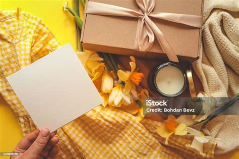 수선화의 아늑한 봄 배경이 있는 빈 사각형 카드 선물 상자 불이 켜지지 않은 양초 부드러운 니트 패브릭을 들고 있는 손으로 인사말이나 초대장 모형에 적합합니다 꽃 꽃의