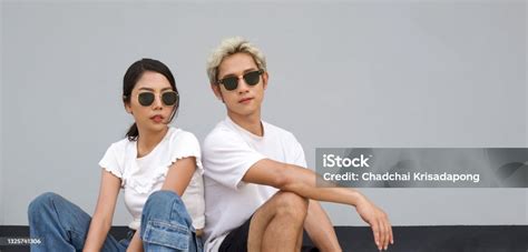 흰색 티셔츠와 검은 색 선글라스를 입은 젊은 아시아 남자가 무릎을 꿇고 팔을 쉬고 있다 그의 여자 친구는 그 옆에 앉아 회색 배경의 세로 아시아에 대한 스톡 사진 및 기타