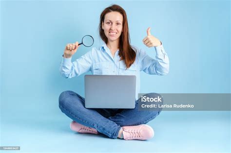인터넷에서 온라인 쇼핑 제품 검색 젊은 성인 소녀가 그녀의 무릎에 노트북과 함께 앉아 파란색 배경에 그녀의 손에 돋보기를 들고