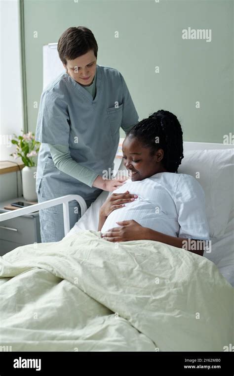 Nurse Wearing Scrubs Assisting Pregnant Patient In Hospital Bed Being