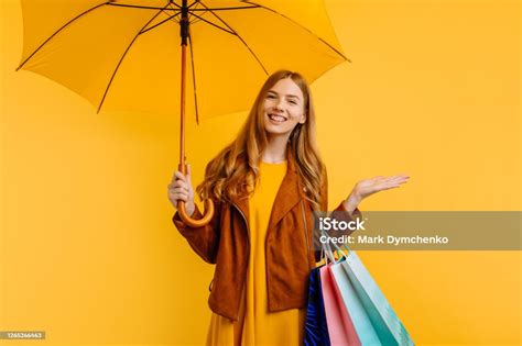 밝은 노란색 드레스와 가을 재킷을 입고 노란 우산과 쇼핑백을 들고 고립된 노란색 배경에 서있는 매력적인 소녀 쇼핑 컨셉 가방에