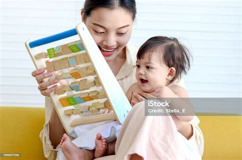 행복한 미소를 짓는 아기는 거실의 노란색 소파에 엄마 무릎에 앉아 있고 작고 귀여운 소녀는 발달 장난감을 가지고 놀고 엄마는 딸