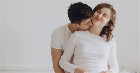 하얀 침대에 배꼽 범프에 하얀 작은 양말을 들고 있는 행복한 젊은 임신한 부부 세련된 임산부 가족 엄마와 아빠는 흰색으로