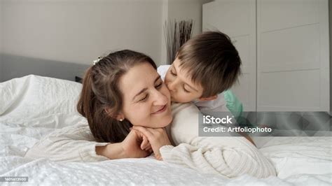 잠옷을 입은 귀여운 소년이 침대에서 어머니와 함께 누워 키스를 하고 있다 가족 행복의 개념 집에서 휴식을 취하고 침대에서 즐거운 시간을 보내고 부모와 쾌활한 아이들 어머니에