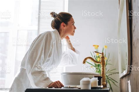 집에서 욕실에있는 거울 근처의 흰색 목욕 가운에 어두운 긴 머리카락을 가진 젊은 여성 불타는 듯한에 대한 스톡 사진 및 기타