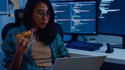 Young Asian Woman Software Developers Eating Burgers And Using Computer To Write Code Sitting At