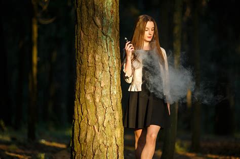 Vape 십대 캐주얼 한 옷을 입은 어린 귀여운 소녀는 여름에 일몰에 숲에서 야외에서 전자 담배를 피울 수 있습니다 나쁜 습관
