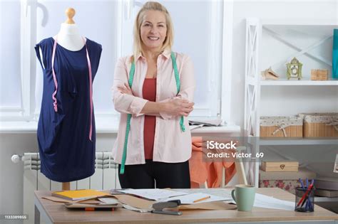 성공적인 여성 디자이너 옷 전체 성장에 스튜디오에서 재단사 아름 다운 유럽 여자 카메라를 보고 배경에 매달려 옷과 워크샵에서 테이블 근처에 서 고객에 대한 스톡 사진 및