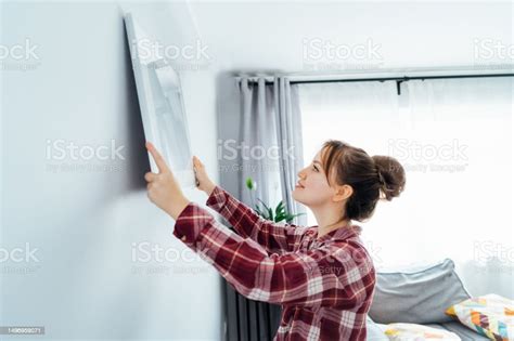 옆모습은 젊은 여자가 벽에 그림을 그리고 모형 액자를 놓는다 캐주얼한 아가씨는 새로운 세련된 아파트에서 아늑함을 돌보고 있다