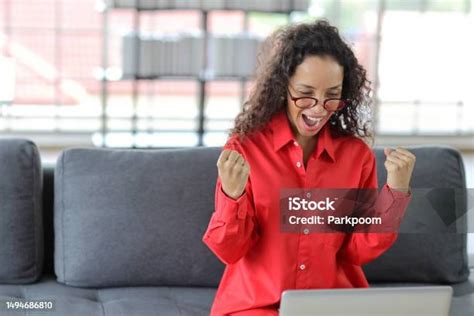 행복한 여자는 앉아서 컴퓨터를 사용하고 행복한 웃는 얼굴로 손을 들고 거실에서 사업 거래를 마쳤다 성공한 무언가로 손을 들고 흥분하는 감정 사업가 25 29세에 대한 스톡