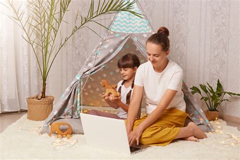 백인 여성과 그녀의 딸이 노트북 앞에서 가발왈에 앉아있는 흰색 티셔츠를 입고 있고 나무 장난감을 가지고 노는 어린 아이