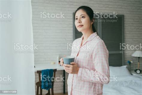 잠옷을 입은 아시아 여성이 창문 근처에 서서 침실에서 일어나고 나서 화창한 아침과 차를 즐기면서 거리를 바라보고 있습니다 건강한